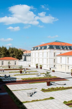 Pátio com um edifício grande, com telhado de telhas vermelhas e paredes brancas. Há plantas em vasos e áreas de estar.