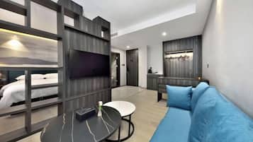 Quarto de hotel moderno com cama, televisão, sofá, mesinha e cozinha compacta.
