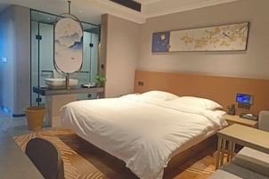 Quarto de hotel com cama grande, mesa de cabeceira, escrivaninha, cadeira, espelho e uma obra de arte pendurada na parede.