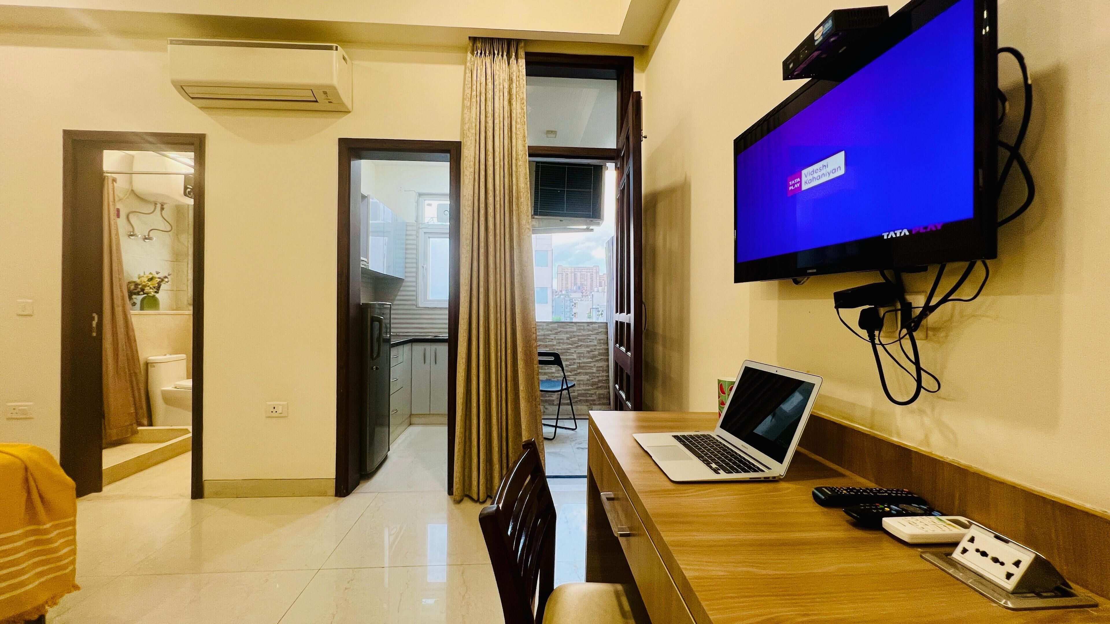 Quarto de hotel com televisão de tela plana na parede, um laptop sobre uma mesa de madeira e uma varanda com vista para a cidade.
