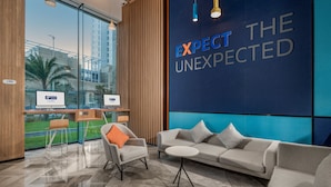 Um lobby de hotel moderno com uma grande janela, uma mesa de recepção, um sofá e uma parede com o texto "EXPECT THE UNEXPECTED".