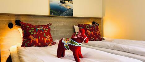 Quarto de hotel com duas camas, cada uma com roupa de cama branca e almofadas vermelhas estampadas. Há uma cabeceira de madeira, uma luminária na parede e um quadro emoldurado sobre as camas.