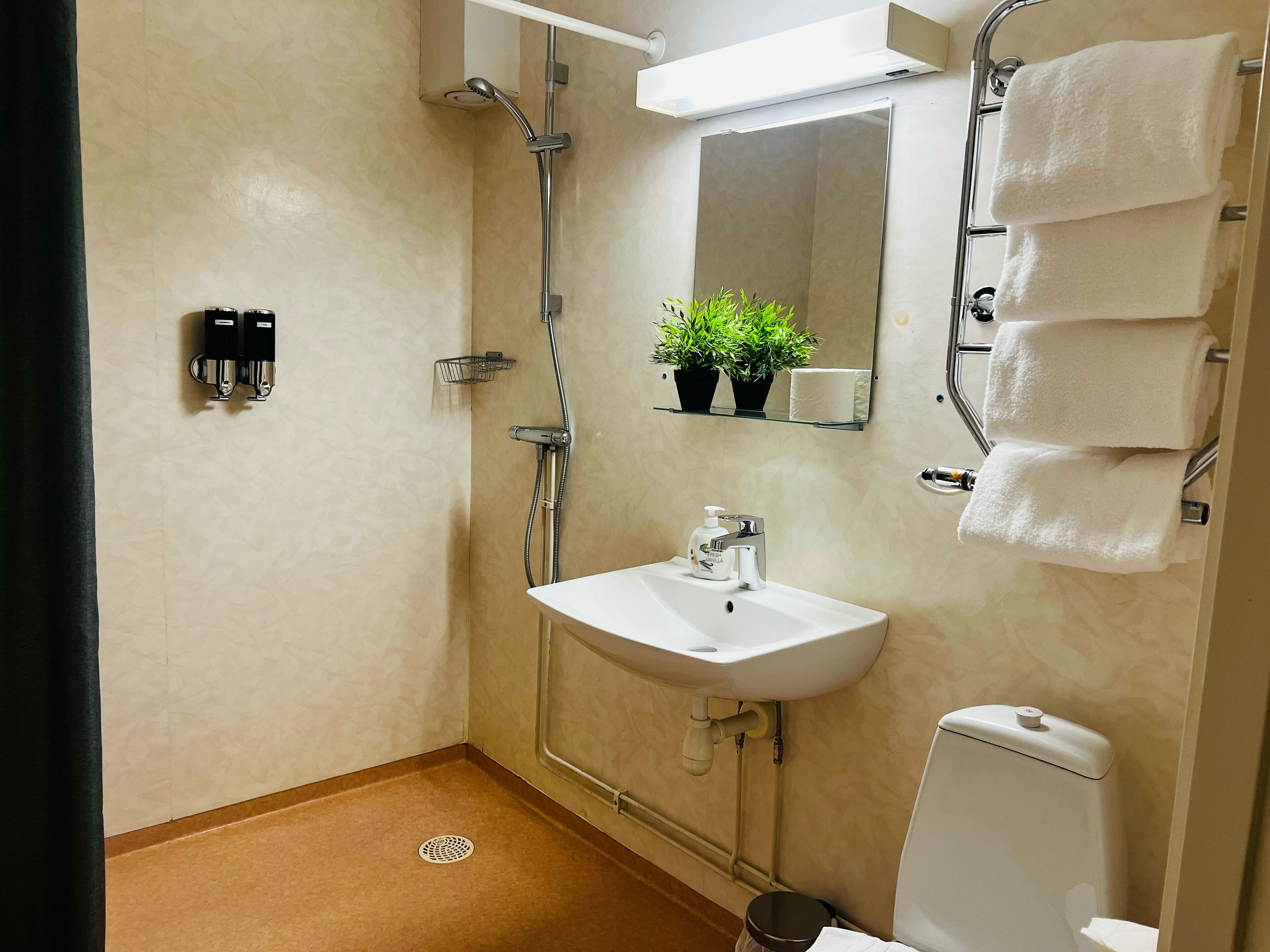 Um banheiro moderno com pia branca, espelho, vaso sanitário e chuveiro. Há duas plantas em vasos na parede e um porta-toalhas com toalhas.