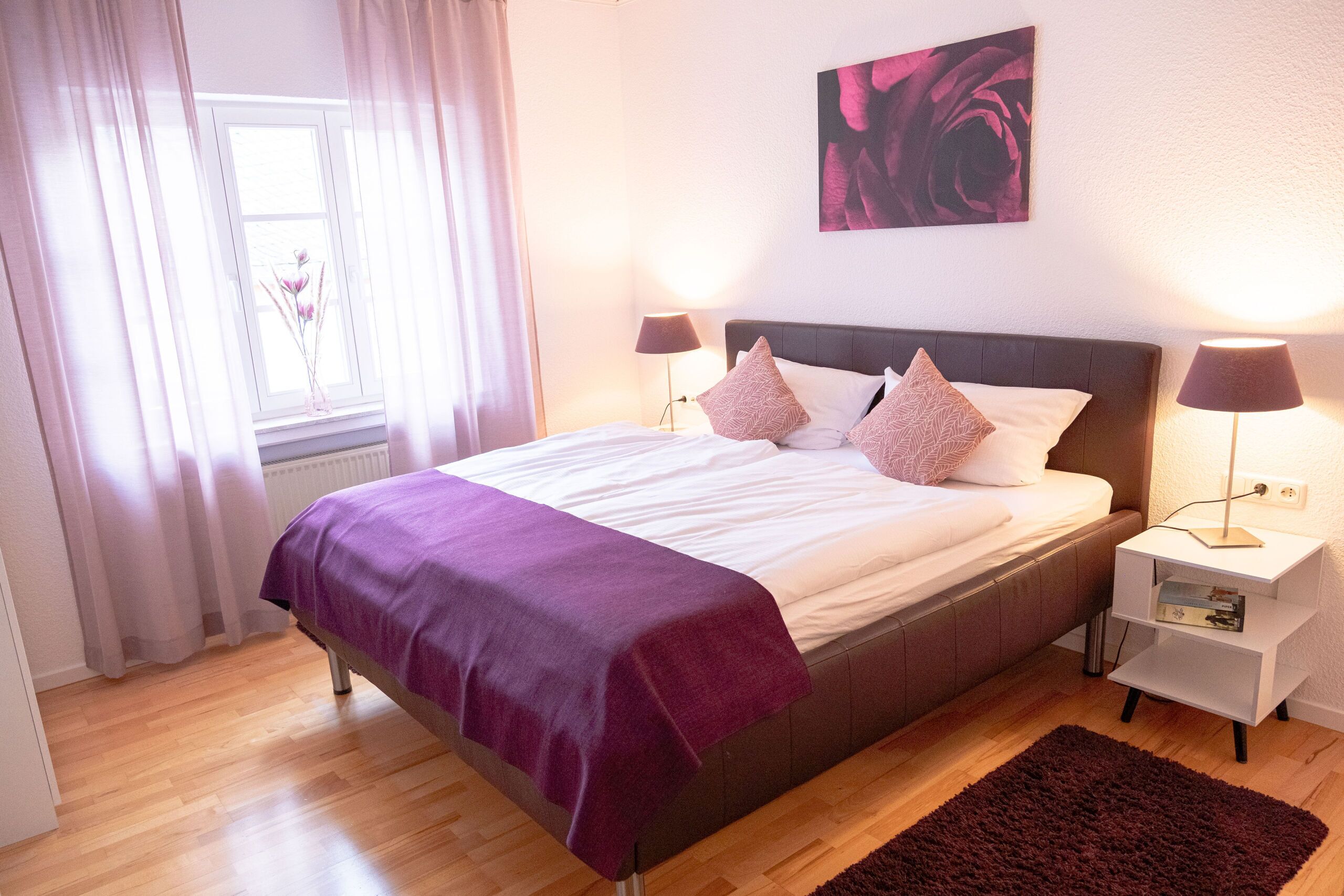 Quarto com cama, duas luminárias de cabeceira, mesa de cabeceira, janela com cortinas e uma obra de arte na parede.