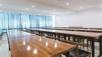 Sala de conferências com várias mesas e cadeiras, amplas janelas com cortinas e teto branco com iluminação embutida.