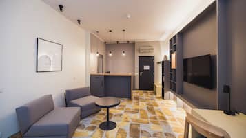 Quarto de hotel moderno com sofá, uma mesinha redonda, televisão e cozinha compacta com bancada e armários.