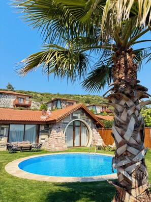 Piscina rodeada por uma casa de pedra com telhado de telhas, uma palmeira e céu azul e límpido.