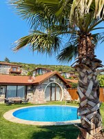 Piscina rodeada por uma casa de pedra com telhado de telhas, uma palmeira e céu azul e límpido.