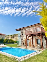 Um edifício de pedra com piscina, deck de madeira e vista para uma paisagem montanhosa.