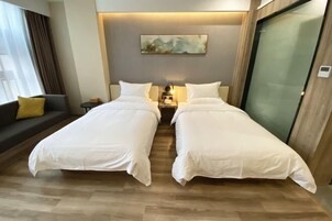 Quarto de hotel com duas camas de solteiro, uma mesinha com abajur, um sofá com almofada amarela e um quadro na parede.