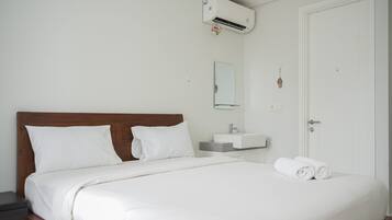 Cama bem arrumada com lençóis brancos, cabeceira de madeira, mesa de cabeceira, ar condicionado branco fixado na parede, espelho pequeno ao lado da cama e uma toalha sobre a cama.