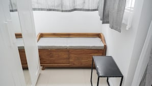 Quarto pequeno e limpo, com um banco de madeira, uma mesa pequena e piso branco.