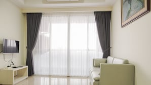 Uma sala de estar moderna com um sofá, uma TV sobre um móvel e grandes janelas com cortinas leves.