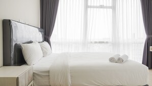 Cama bem arrumada com lençóis brancos e duas toalhas enroladas, uma mesinha de cabeceira e uma janela grande com cortinas finas.