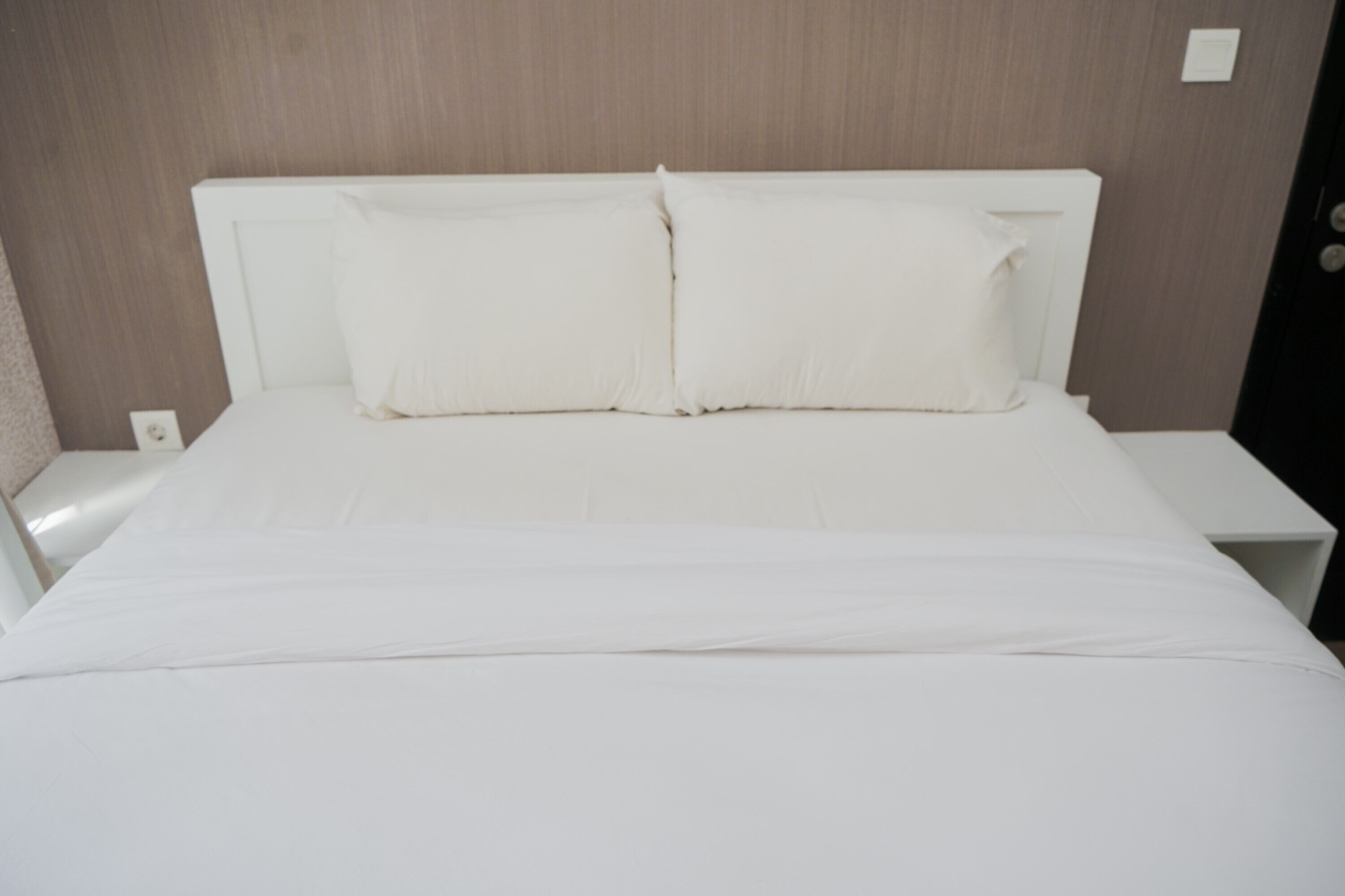 Cama bem arrumada com lençóis e almofadas brancas, cabeceira de madeira e mesa de cabeceira branca com tomada.
