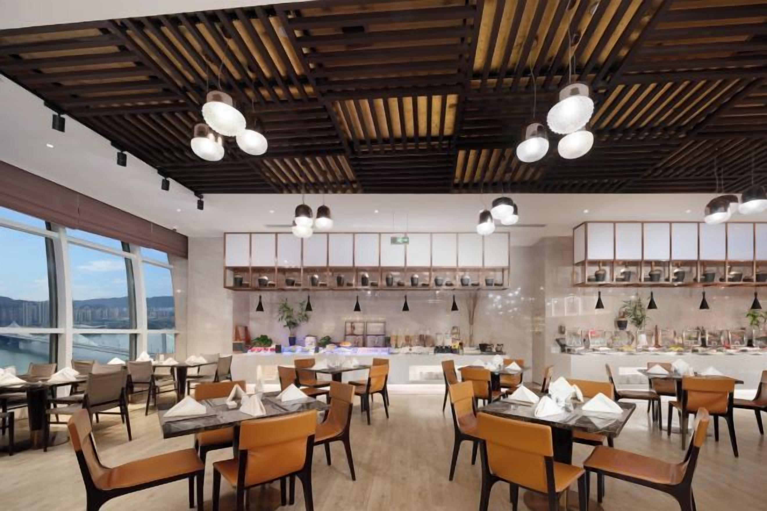 Um restaurante espaçoso com pé direito alto, luminárias modernas e vista para a cidade.