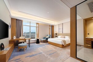 Quarto de hotel com uma cama grande, uma escrivaninha com cadeira, televisão e vista para a cidade.