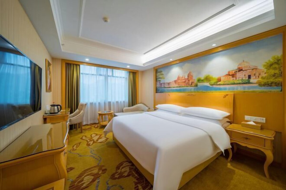 Quarto de hotel com uma cama grande, televisão de tela plana, escrivaninha com cadeira, mesa redonda pequena e um quadro de um edifício histórico na parede.