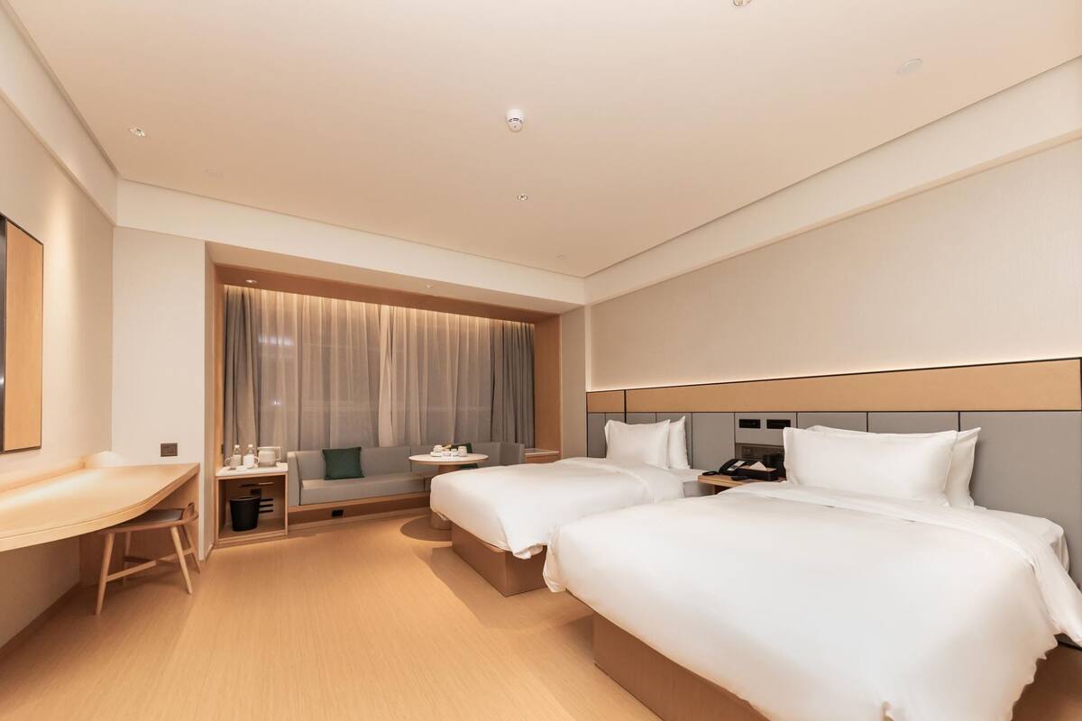 Quarto de hotel com duas camas, uma escrivaninha, um sofá e uma janela grande com cortinas.