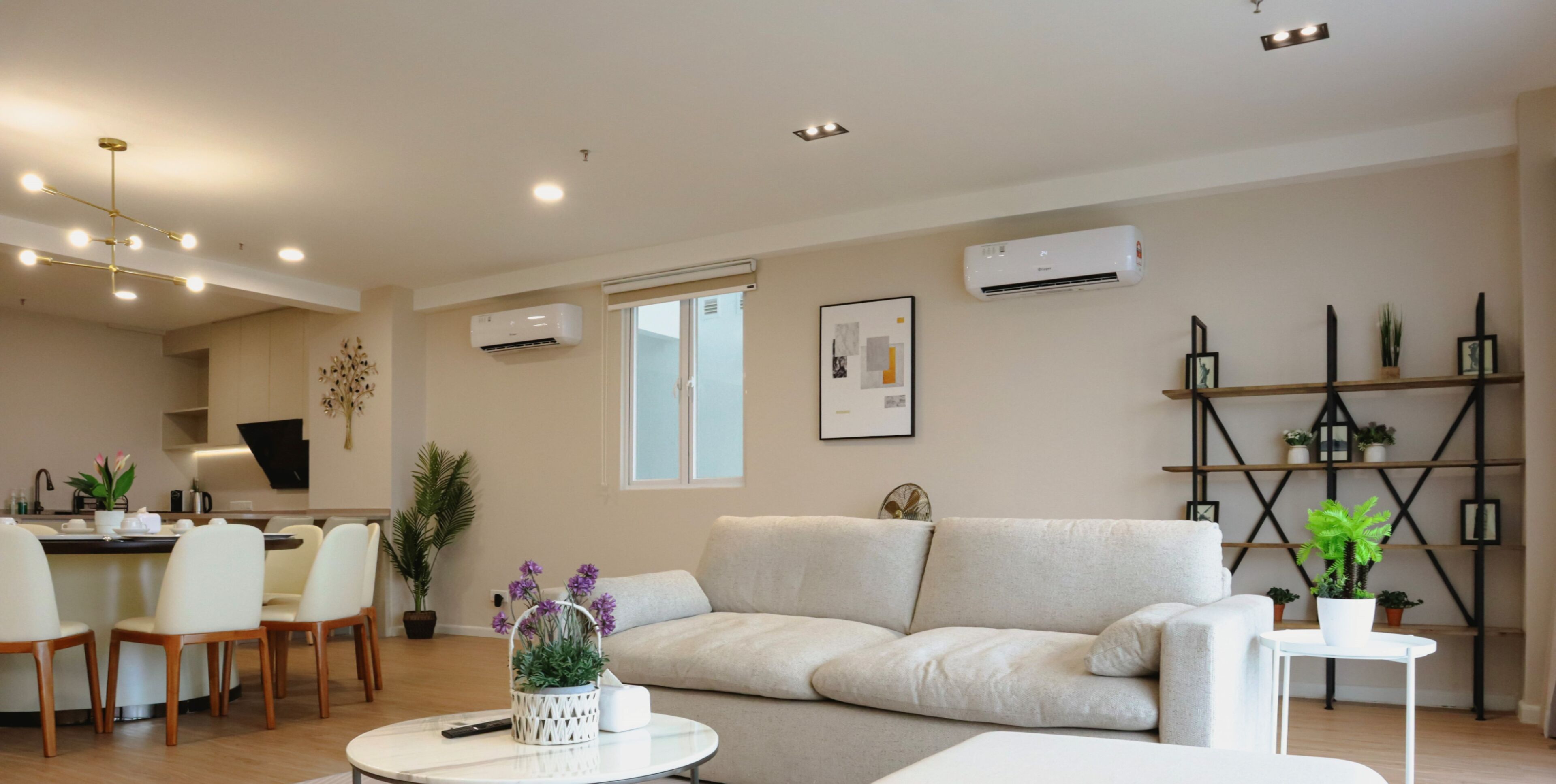 Uma sala de estar moderna com sofá, área de jantar e cozinha. Há unidades de ar condicionado na parede, um lustre e uma estante com plantas.