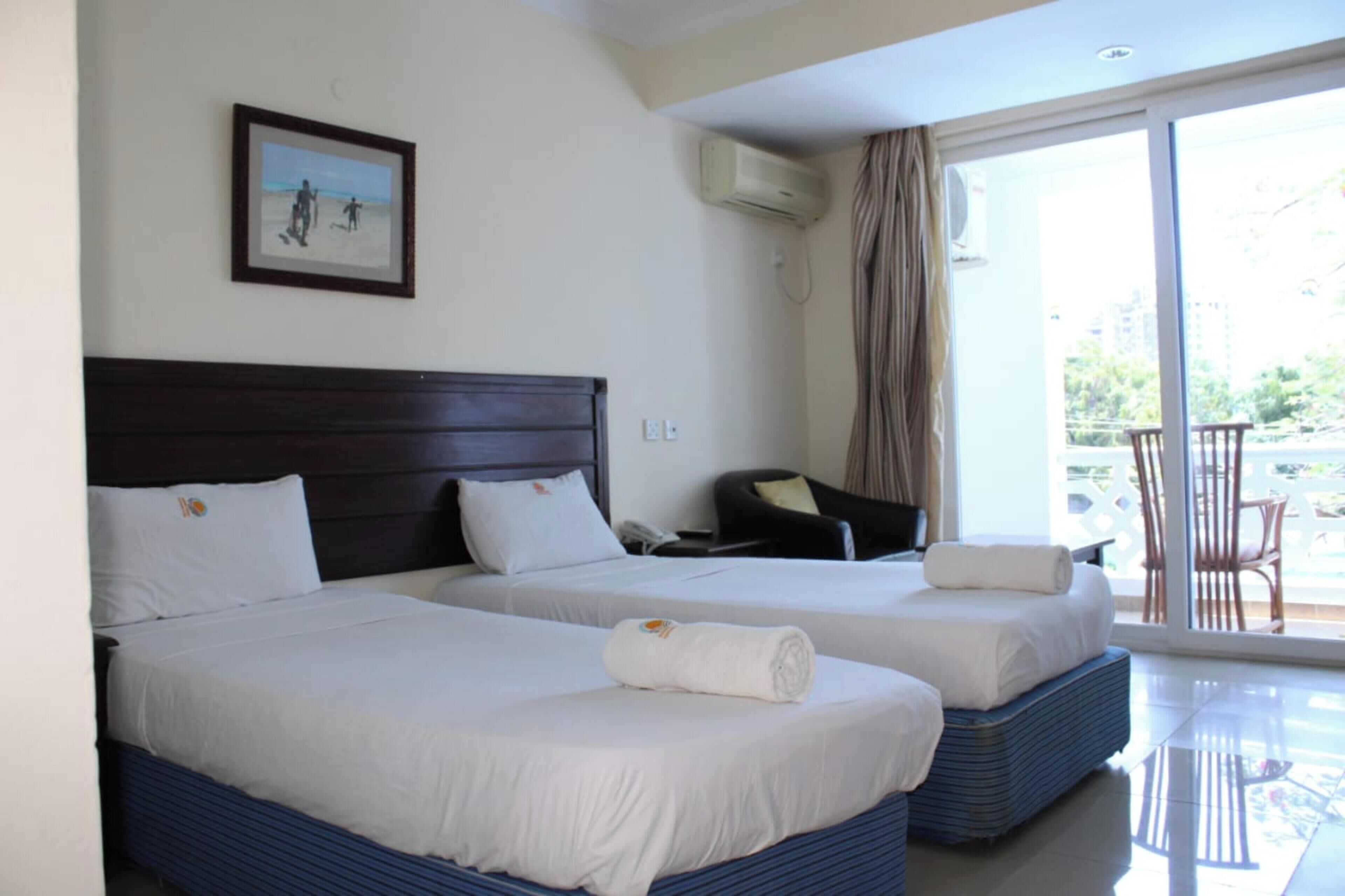 Quarto de hotel com duas camas, uma cadeira, uma mesa pequena e varanda com cadeiras.