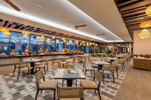 Uma sala de jantar ampla com várias mesas e cadeiras, ventiladores no teto e grandes janelas.
