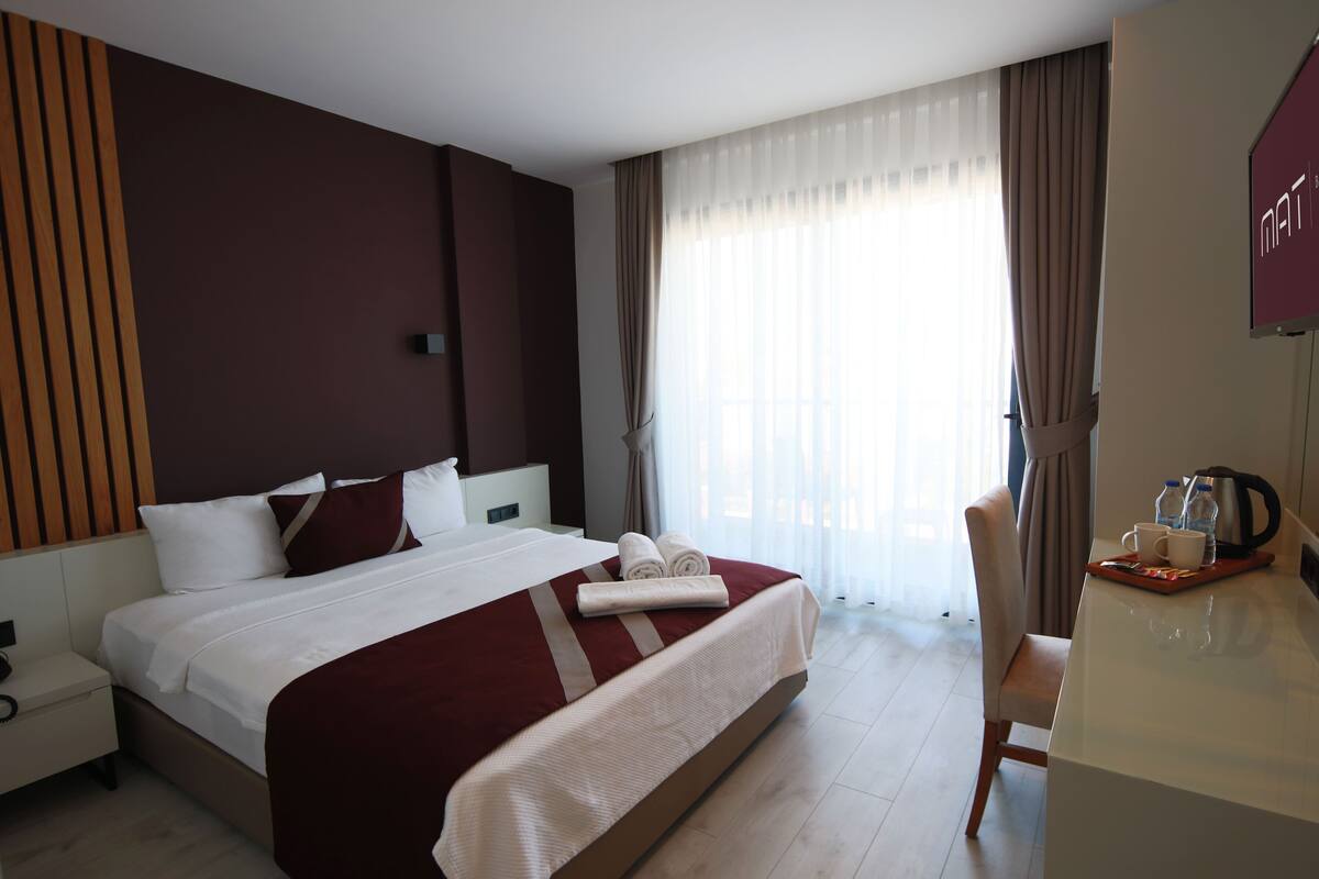Quarto de hotel com uma cama grande, uma mesa com chaleira e uma cadeira.