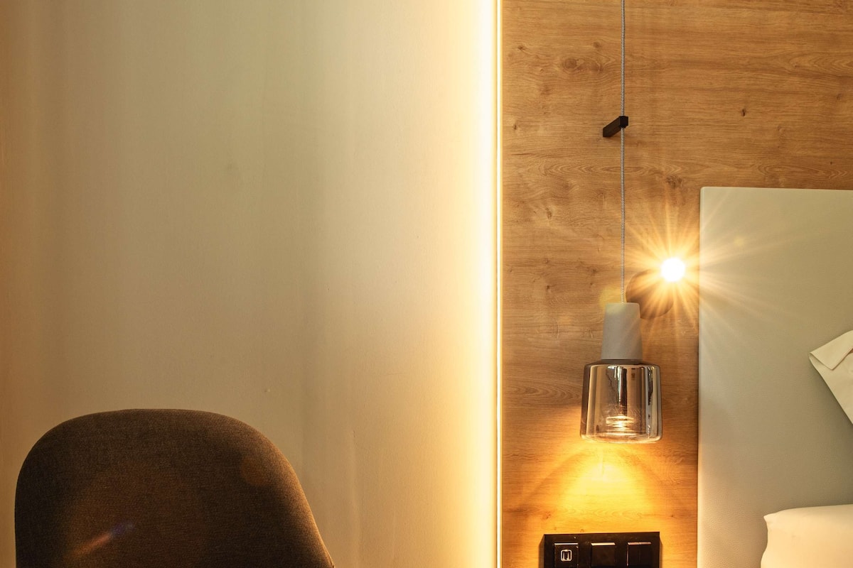 Quarto moderno com parede de destaque em madeira, luminária de chão, mesa lateral redonda e poltrona estofada escura.