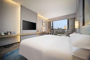 Quarto de hotel moderno com uma cama grande, televisão de tela plana, escrivaninha com luminária e vista para a paisagem urbana.