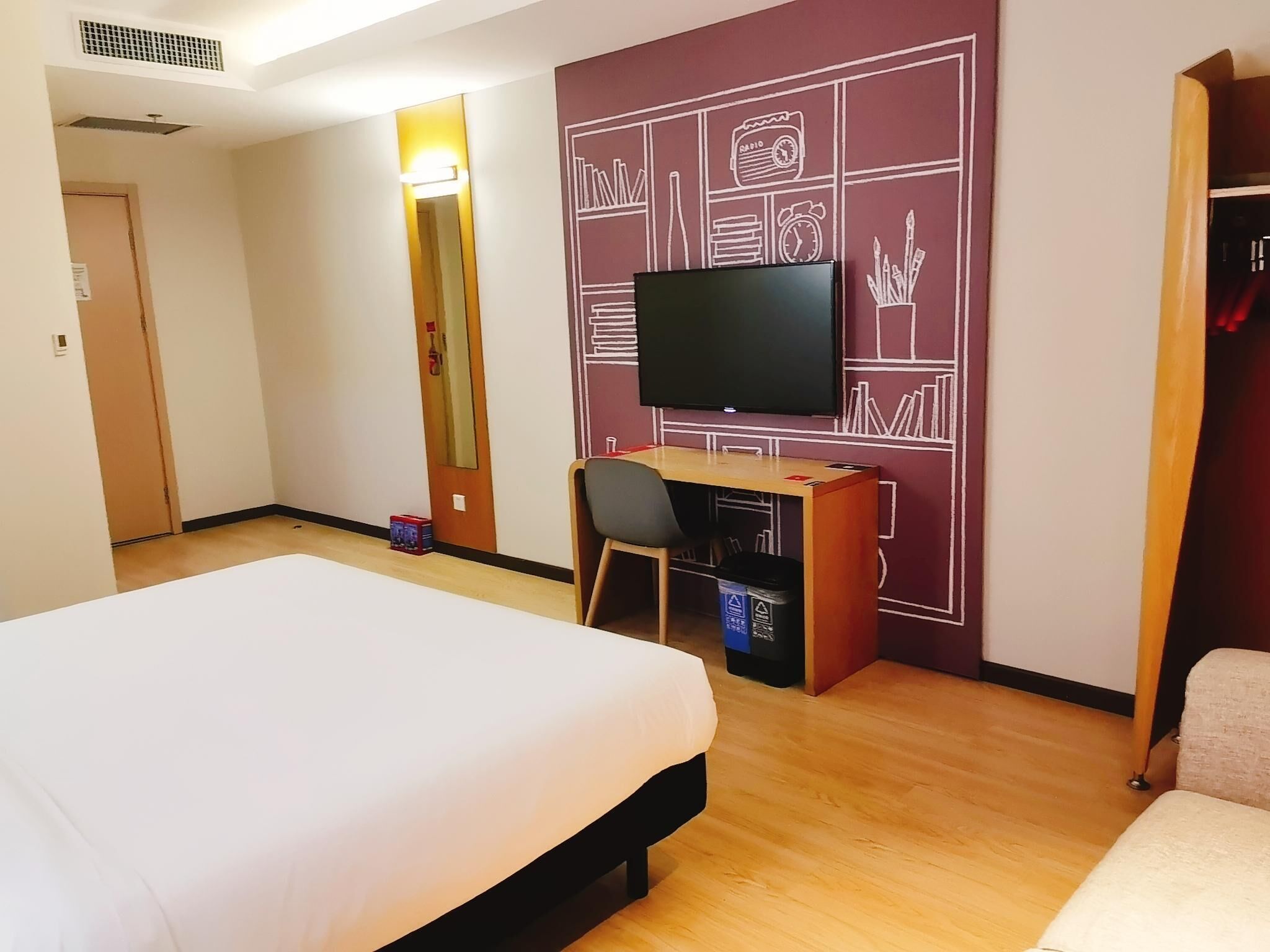 Quarto de hotel com cama, escrivaninha e cadeira, televisão e parede com design geométrico.