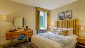 Quarto de hotel com cama, escrivaninha com espelho, cadeira, janela com cortinas e mesa de cabeceira com abajur.