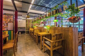 Um restaurante com mesas e cadeiras de madeira, um espaço de bar com plantas penduradas e vista pela janela.