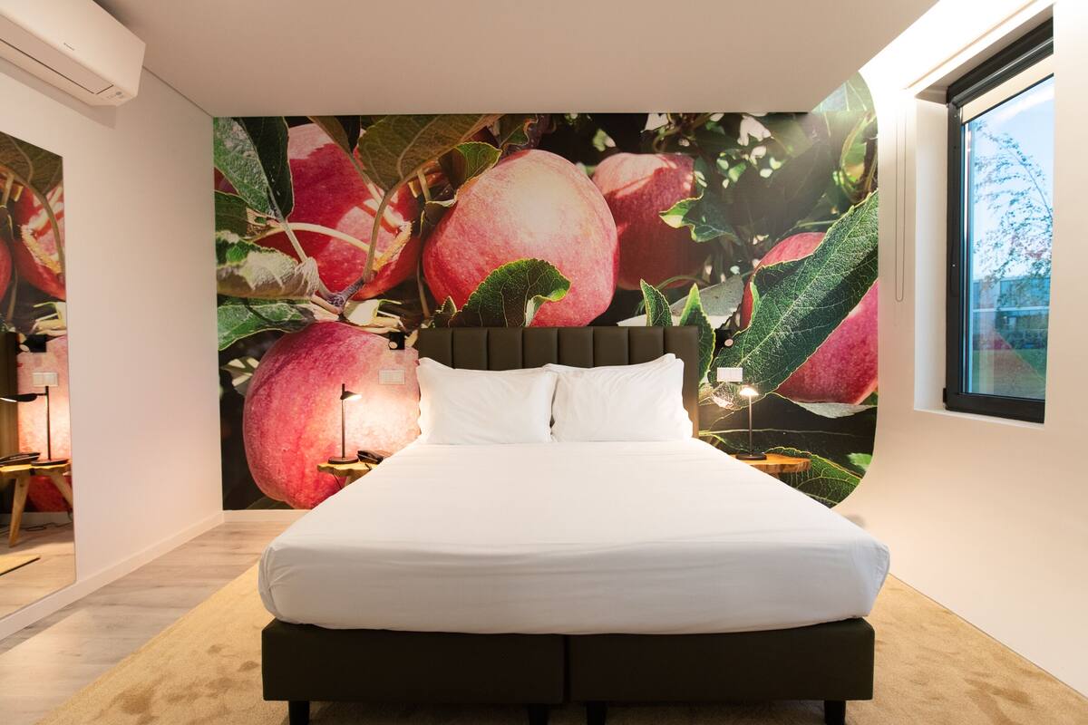 Quarto com cama, painel decorativo de frutas e janela.