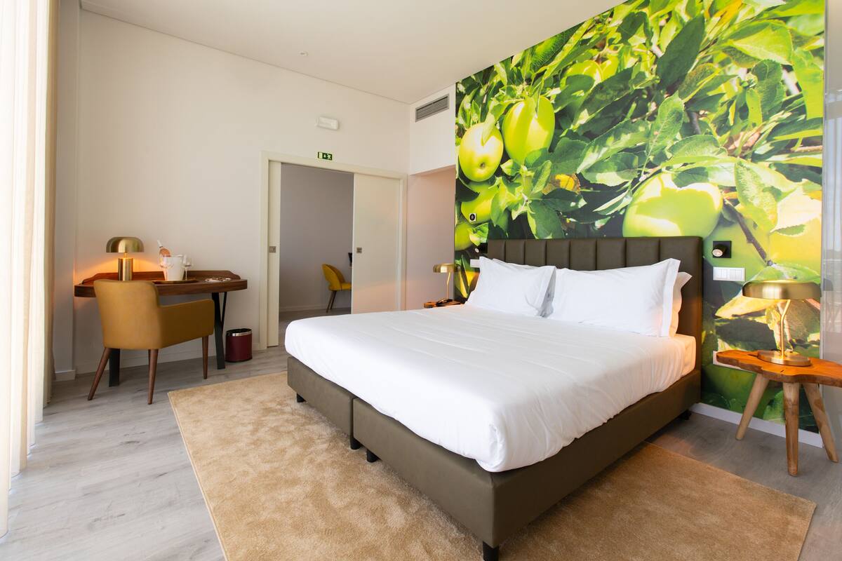 Quarto de hotel com uma cama grande, uma mesa com cadeira, um abajur e um mural na parede.