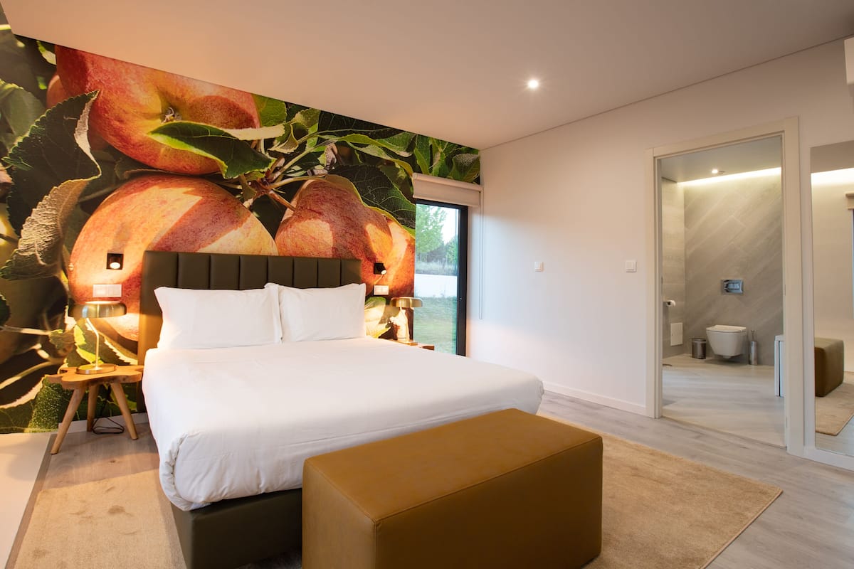 Quarto moderno com uma cama grande, uma mesa de cabeceira de madeira, um mural na parede e vista para o banheiro.