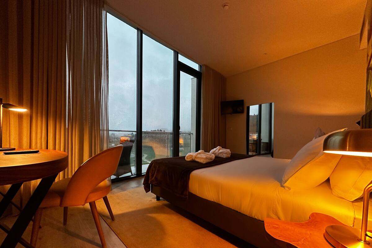 Quarto de hotel com uma cama grande, uma mesa, uma cadeira e vista pela janela.