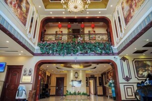 Um saguão de hotel com uma escadaria imponente, uma varanda com vegetação e um lustre.