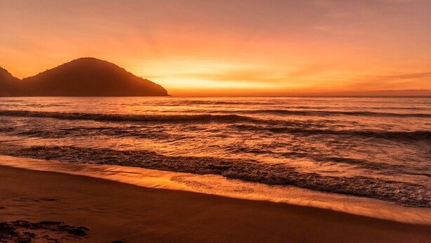 Uma praia com ondas ao pôr do sol, uma montanha distante e céu claro.