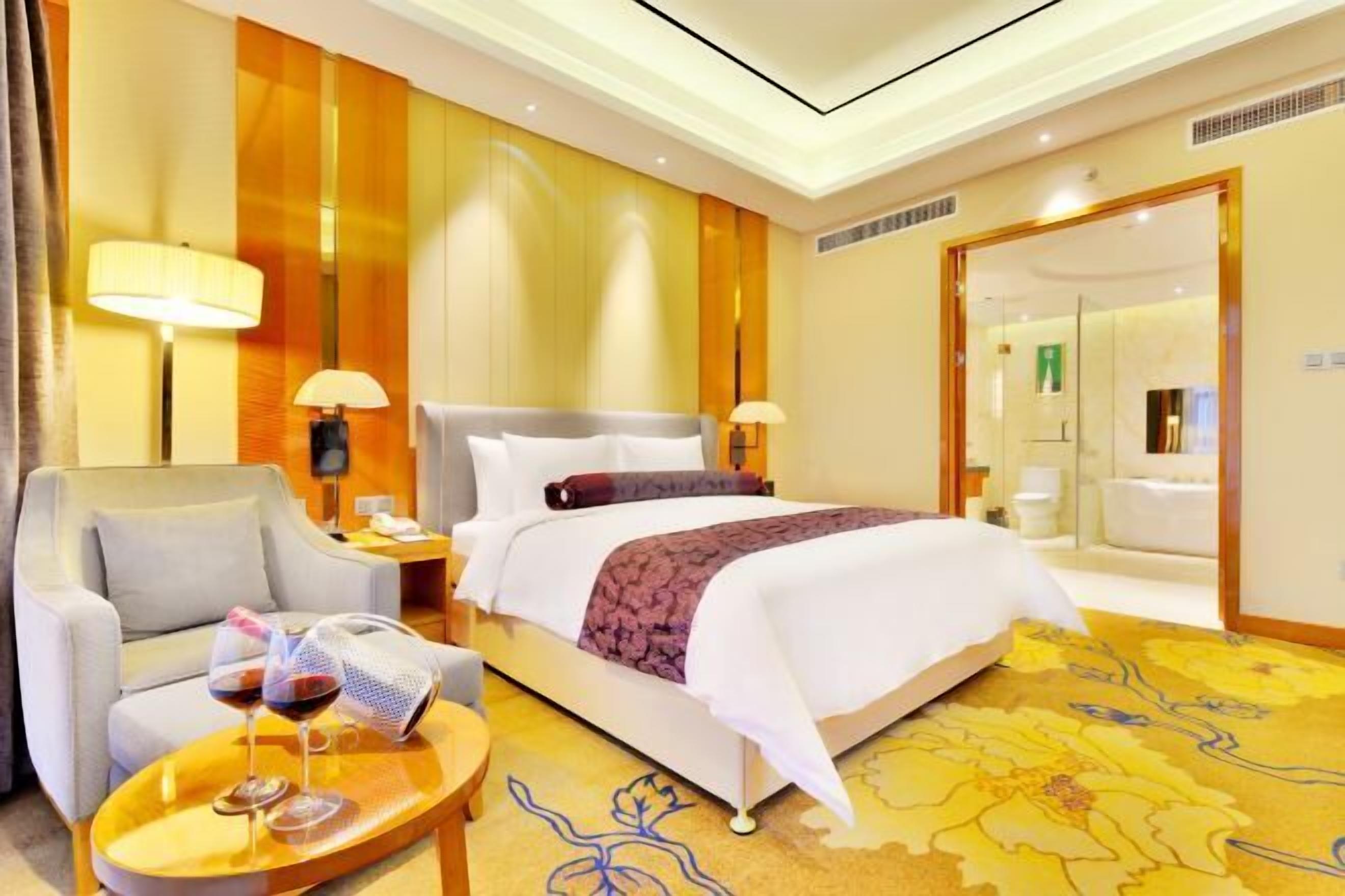 Quarto de hotel com uma cama, um sofá, uma mesinha com copos de vinho e um banheiro visível por uma porta aberta.