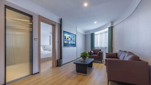 Quarto de hotel moderno com piso de madeira, um sofá marrom, uma mesinha com uma planta, uma cama com lençóis brancos e um quadro na parede.