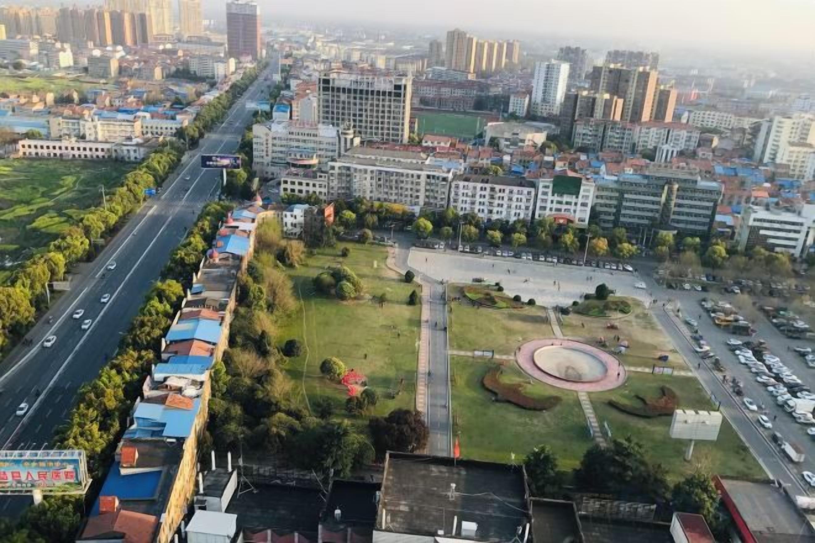 Paisagem urbana com um grande complexo de edifícios, um cruzamento rotatório e uma estrada arborizada.