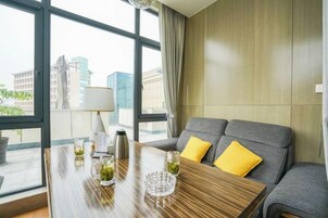 Quarto de hotel com sofá, mesa de madeira e vista para prédios através de grandes janelas.