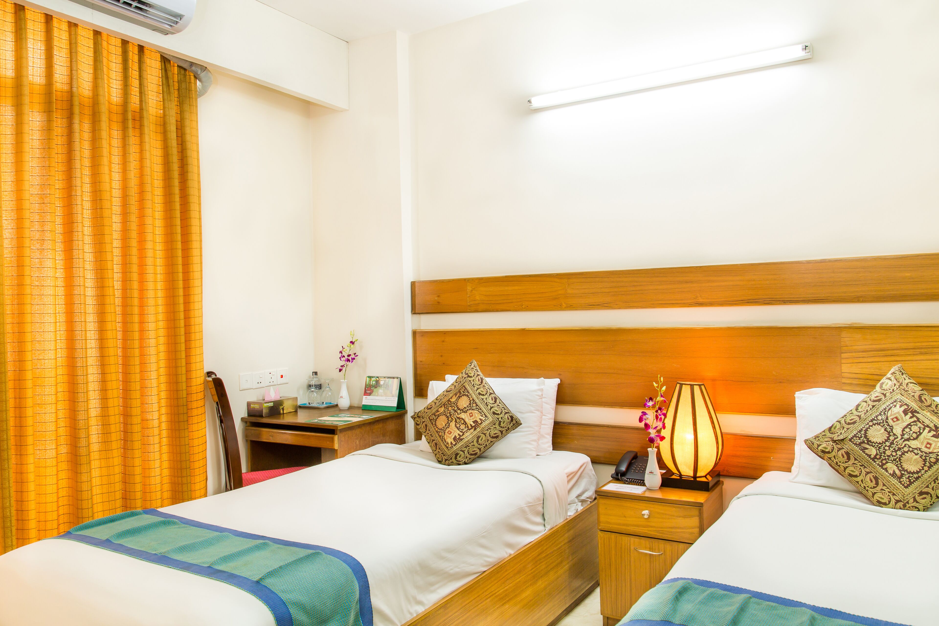 Quarto de hotel com duas camas, cabeceira de madeira, mesas de cabeceira com luminárias, uma escrivaninha e uma cadeira.