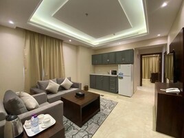 Uma sala de estar moderna com um sofá, mesa de centro e uma bandeja com chá e chaleira. Uma cozinha com armários e geladeira é visível ao fundo.