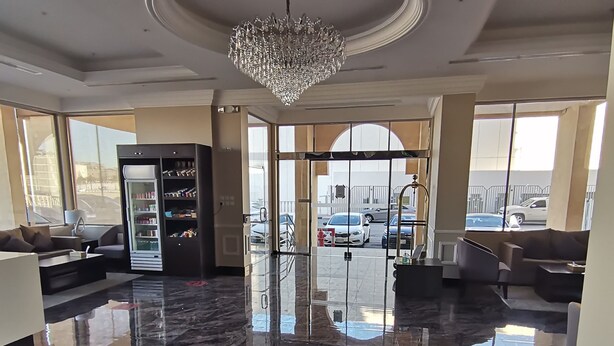 Um hall amplo com piso de mármore, um lustre, balcão de recepção e grandes janelas com vista para carros estacionados lá fora.