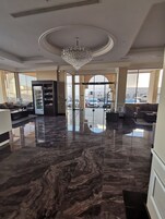 Um hall amplo com piso de mármore, um lustre, balcão de recepção e grandes janelas com vista para carros estacionados lá fora.