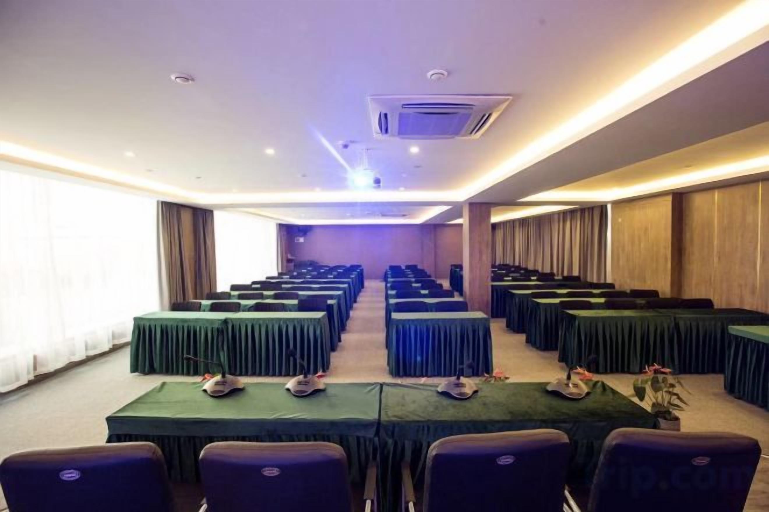 Uma sala de conferência com fileiras de mesas e cadeiras, um palco na frente e janelas amplas em uma lateral.