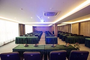 Uma sala de conferência com fileiras de mesas e cadeiras, um palco na frente e janelas amplas em uma lateral.