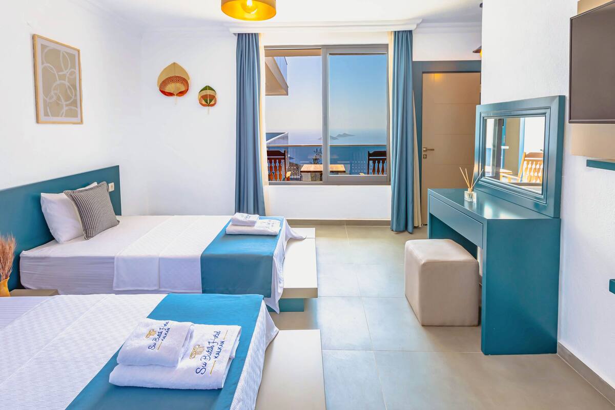 Quarto de hotel com duas camas, uma escrivaninha, uma cadeira, varanda com vista e um espelho grande.