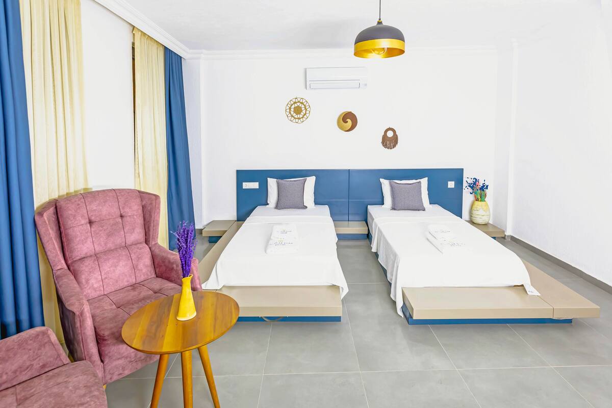 Quarto de hotel com duas camas de solteiro, uma poltrona rosa, uma mesinha de madeira com um vaso, e uma luminária pendente moderna.